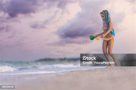 여름 의상을 입은 아프리카 머리띠를 가진 소녀가 화창한 일몰 아래 파도가 있는 바다 근처의 조개와 함께 해변에서 놀고 있습니다 땋은 머리에 대한 스톡 사진 및 기타 이미지