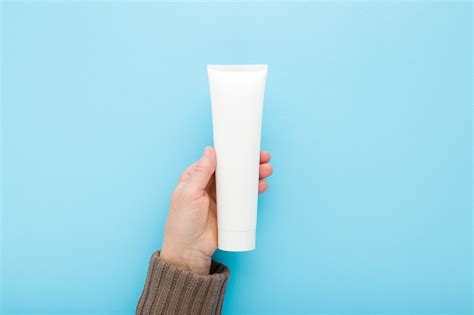 성숙한 여자 손 잡고 밝은 파란색 배경에 안티 에이징 크림의 흰색 튜브를 보여주는 파스텔 색상 근접 촬영 노년기에 깨끗하고 부드러운 체신에 신경을 쓰다 뷰티 제품 하향 보기
