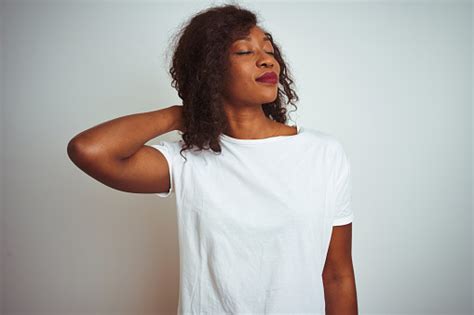 고립된 흰색 배경 위에 서있는 티셔츠를 입은 젊은 아프리카 계 미국인 여성은 목 통증 손으로 목을 만지고 근육통을 앓고 있습니다 T 셔츠에 대한 스톡 사진 및 기타 이미지