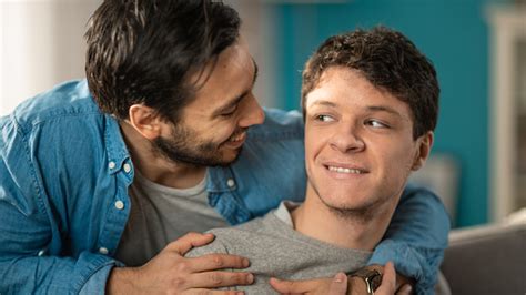 집에서 귀여운 남성 퀴어 커플의 초상화입니다 젊은 남자는 소파에 앉아 그의 파트너는 뒤에서 와서 부드럽게 그를 포용 그들은 행복하고 웃고 있습니다 객실은 현대적인 인테리어로