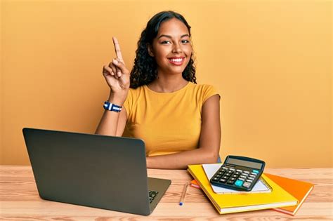 노트북과 계산기로 사무실에서 일하는 젊은 아프리카 계 미국인 소녀는 성공적인 아이디어로 손가락을 가리킵니다 빠져나와 행복했다 번호 1 계산기에 대한 스톡 사진 및 기타