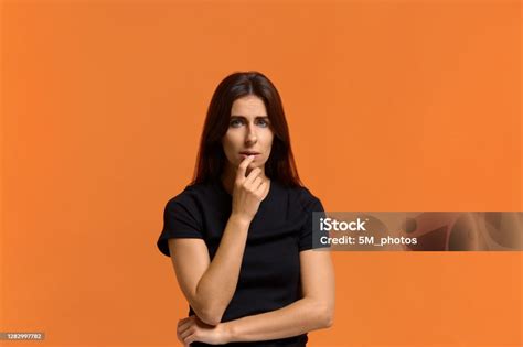 어떻게 생각 그것은 좋은 생각이다 검은 색 티셔츠를 입은 주저하는 백인 여성의 이미지가 입술에 집게 손가락을 들고 무엇을 해야할지 의심합니다 주황색 배경 위에 격리됩니다 T