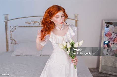 귀에 하얀 진주 귀걸이와 흰색 빈티지 웨딩 드레스에 긴 붉은 곱슬 머리 여자 창백한 피부 파란 눈 손에 흰색 튤립으로 침실에