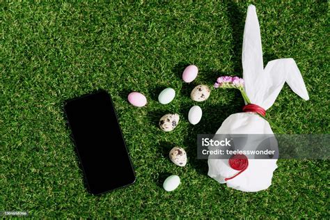 초록색 잔디 배경 위에 파스텔 색칠과 메추라기 달걀로 유약 된 초콜릿 부활절 달걀 토끼 모양의 흰 베이킹 페이퍼 검은 휴대 전화 행복한 부활절 개념 공간을 복사합니다 위쪽