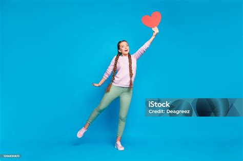 머리띠 피그 테일을 가진 쾌활한 청소년 소녀의 전체 길이 사진은 14 월 핑크 스웨터 녹색 세련된 바지 고립 된 파란색 배경에 손을 잡는 비행 종이 카드 심장 놀라움을 잡고