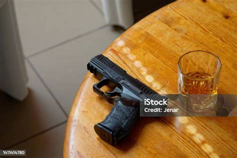나무 테이블에 검은 손총이 열리고 흐릿한 문이 바닥에 드리워진 그림자가 배경에 있습니다 위스키가있는 유리가 무기 근처에 머물러 있습니다 0명에 대한 스톡 사진 및 기타