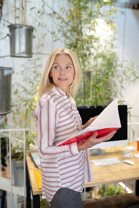 Blonde Woman Standing At Desk Holding Notebook Looking Sideways Stock