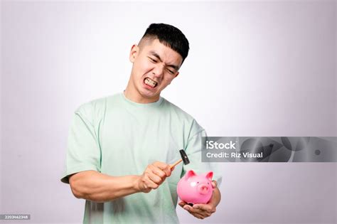 민트 그린 셔츠를 입은 좌절한 아시아 남자가 금융 위기 비상 저축을 상징하는 흰색 배경에 작은 망치와 분홍색 돼지 저금통을 들고 얼굴을 찡그리고 있습니다 30 34세에 대한