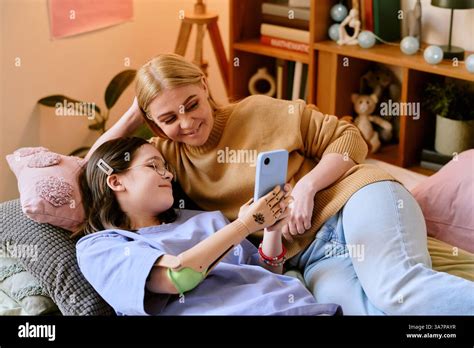 Blonde Mom And Her Brunette Daughter With Prosthetic Hand Sitting On Sofa With One Holding