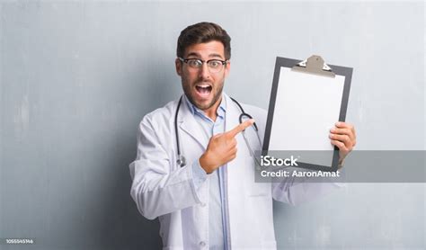 잘생긴 젊은 매우 행복 손과 손가락으로 가리키는 클립보드를 들고 회색 그런 지 벽에 남자 의사 가리키기에 대한 스톡 사진 및 기타 이미지 Istock