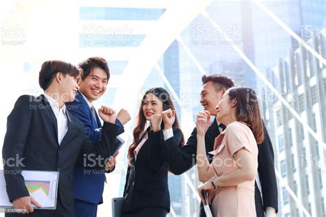 성공을 축하하기 위해 주먹을 쥔 5명의 젊은 아시아 쾌활한 사업가 그룹 사업가와 여성은 고층 빌딩이 흐릿한 배경으로 외부에서 강력한 팀워크를 약속하며 동료들을 응원합니다