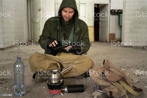 더러운 피곤한 남자가 버려진 방의 바닥에 앉아 가스 버너 위로 손을 따뜻하게하고 저녁 식사를 준비합니다 포스트 묵시록 포스트