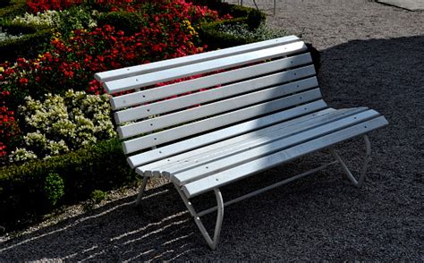 공공 공원에서 금속 빛나는 스테인레스 스틸 구조 팔걸이 흰색 나무 패널벤치 화강암 조약돌 타일의 벤치 아래 하얀 꽃이 피는 다년생공원 잔디밭 조화 강철에 대한 스톡 사진 및