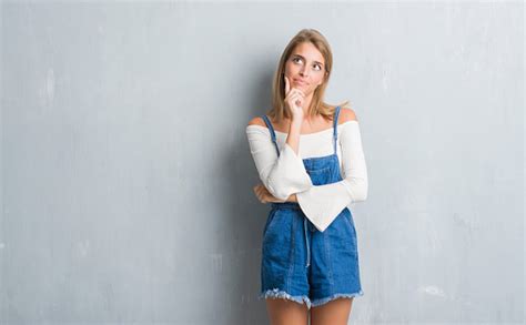 손으로 턱 질문 잠겨있는 표현에 대 한 생각에 그런 지 회색 벽에 서 있는 아름 다운 젊은 여자 사려깊은 얼굴 미소 의심 개념입니다 낙하복에 대한 스톡 사진 및 기타 이미지