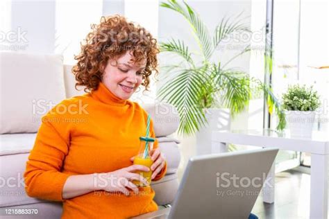 귀여운 여성 빨간 머리 프리랜서 독서 노트북 동안 현대 가정에서 열린 컴퓨터와 바닥에 앉아 오렌지 주스를 하면서 노트북에서 일하는 젊은 창조적 인 여자 곱슬 머리에 대한