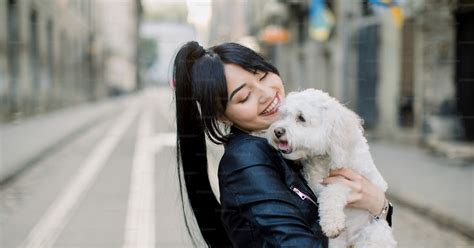 검은 가죽 재킷을 입은 젊은 검은 머리 아시아 여성이 오래된 유럽 도시 거리의 거리에 서서 하얀 작은 개를 안고 포옹합니다 수평 도시 샷 복사 공간 사진