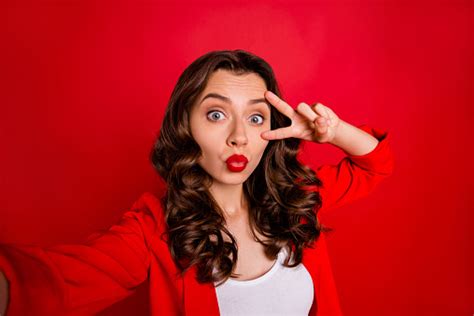 빨간 배경 위에 고립 된 블레이저 재킷을 입고 셀카를 찍는 공기 키스를 보내는 V 표지판을 만드는 꿈꾸는 아가씨의 사진을 클로즈업하십시오 갈색 머리에 대한 스톡 사진 및