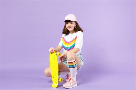 노란 스케이트 보드를 들고 앉아 생생한 옷에 웃는 십 대 소녀 스튜디오에서 보라색 파스텔 벽 배경에 고립 된 카메라를 찾고 사람들은 진심으로 감정 라이프 스타일 개념입니다