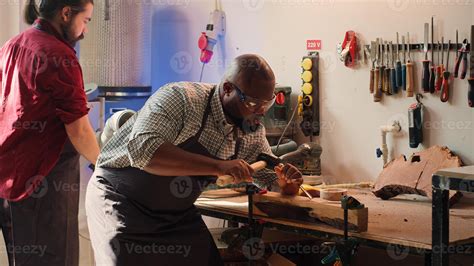 Sculptor Shaping Raw Timber Using Chisel And Hammer In Carpentry Shop With Coworker Cleaning