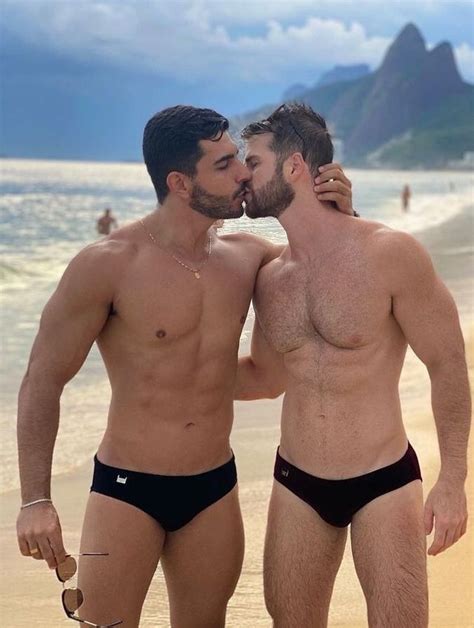 Two Men In Swimsuits Standing On The Beach With Their Heads Close To