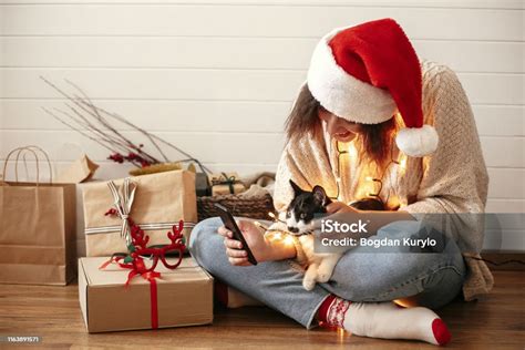 축제 크리스마스 조명과 선물의 배경에 전화 화면을보고 귀여운 고양이와 산타 모자에 세련된 행복 소녀 젊은 힙스터 여자 포옹 키티
