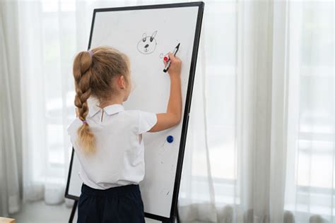 앞 수업에 서서 복사 공간이있는 흰색 보드에 검은 펜으로 그림을 그리는 젊은 백인 예쁜 소녀의 뒷모습 학교에서 수업 중에 자신 만의 스타일을 창조하십시오 어린 시절 교육과