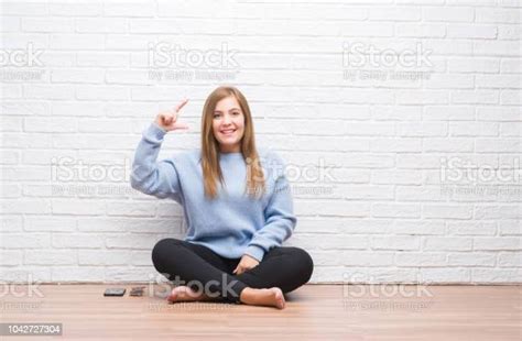 흰색 벽돌 벽 웃 고 자신감 생기고 카메라 동안 손가락으로 크기 표시를 하 고 손으로 몸짓을 통해가 바닥에 앉아 젊은 성인 여자 측정 개념입니다 가을에 대한 스톡 사진 및