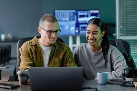 Two Smiling Programmers Cooperating Using Laptop At Desk During Work Meeting In Office Stock