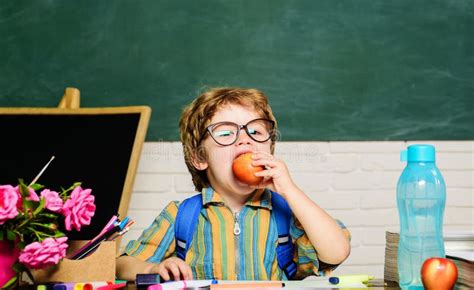 Little Schoolboy In Glasses Sitting At Desk Eating Apple At Lunch Time