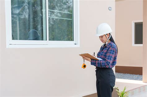 안전모를 쓴 아시아 여성 건물 검사관이 건물 외관을 검사하는 동안 클립보드에 메모를 하고 있습니다 구조적 세부 사항을 확인하고 안전 규정을 보장합니다 옥외 건설 현장 30