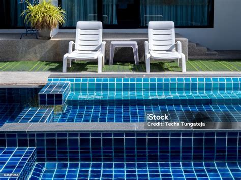 수영장의 녹색 유리에 하얀 선베드를 놓고 벤치 좌석을 띄우고 깨끗하고 맑은 물이 있는 좌석 구역 아무도 없습니다 파란색 타일이 있는 정사각형 모양의 수영장 사람 없음 여름