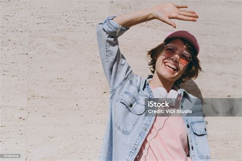 젊은 행복한 학생 백인 여성은 청바지 옷 헤드폰 안경 캡을 입고 도시 거리 야외 산책의 건물 벽에 기대어 태양 사람들 도시 라이프 스타일 개념에서 손으로 커버 거리에 대한