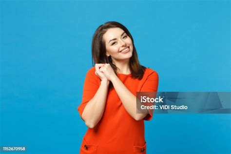 아름 다운 꿈 같은 갈색 머리 젊은 여 자가 파란색 벽 배경 스튜디오 초상화에 고립 된 포즈 서 있는 소원을 만드는 빨간 오렌지 드레스를 입고 사람들의 라이프 스타일 패션