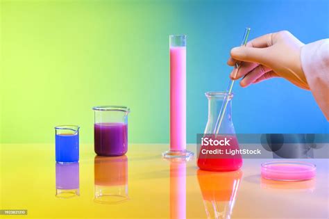 노란색에서 파란색으로 색이 변하는 배경에 알록달록한 액체가 들어 있는 유리 용기 옆에 빨간색 액체가 들어 있는 삼각 플라스크 안에서 유리 막대를 들고 휘젓고 있는 손 개발에