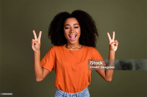 물결 모양의 헤어 스타일을 가진 매우 기뻐하는 쾌활한 소녀의 초상화는 어두운 카키색 배경에 고립 된 V 기호 기호를 보여주는 주황색 티셔츠를 입습니다 T 셔츠에 대한 스톡