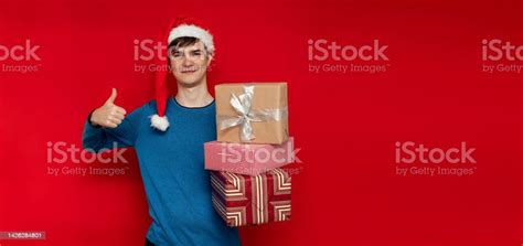 산타 모자를 쓴 청년이 크리스마스 선물을 들고 다른 한 손은 엄지 손가락을 위로 향하게합니다 감정에 대한 스톡 사진 및 기타 이미지 Istock