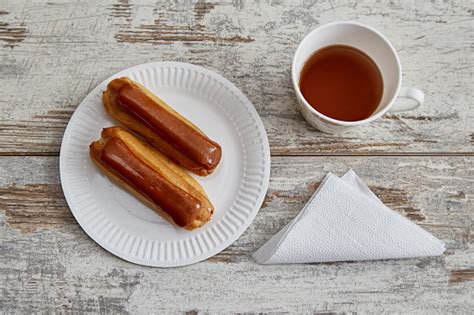 두 개의 Eclairs는 차 한 잔과 냅킨 옆의 나무 탁자 위에 흰 종이 접시에 누워 있으며 클로즈업 섬세한 디저트 고전적인 요리법 수제 크림 패스트리 티타임 낮에 대한