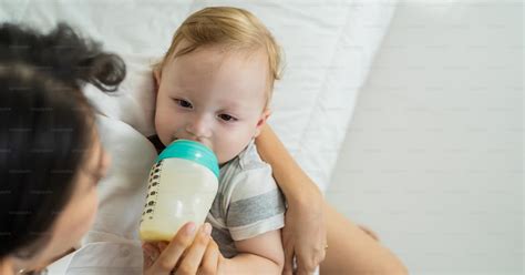 백인 아름 다운 어머니 들고 우유 병에서 아기를 먹이고 먹이는 매력적인 사랑하는 가족 엄마는 침대에 앉아 졸린 작은 유아를 손에 들고 집에서 병에서 아들에게 음료를