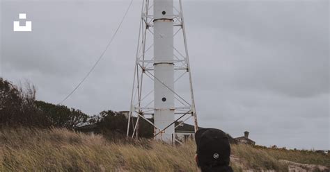 낮 동안 흐린 하늘 아래 흰색과 빨간색 등대 근처에 서 있는 검은 까마귀를 입은 남자 사진 Unsplash의 무료 라트비아 이미지