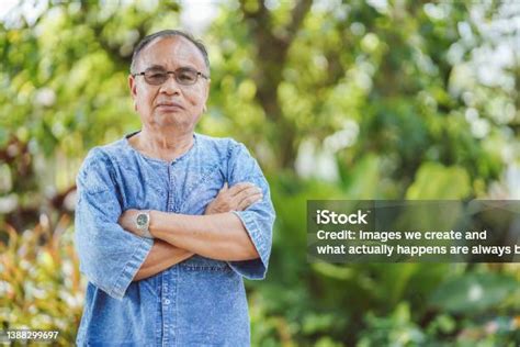 팔을 들고 서 있는 태국 노인이 흐릿한 자연 배경으로 카메라를 바라보며 교차했다 공간을 복사합니다 노인에 대한 스톡 사진 및