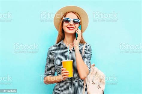 밀짚 모자를 쓰고 주스 한 잔 파란색 배경에 배낭을 들고 스마트폰으로 전화하는 여름 초상화 행복한 미소 여자 거리에 대한 스톡