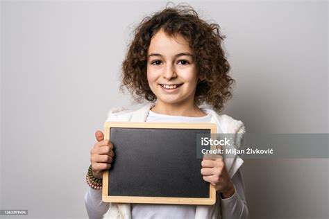 곱슬 머리 앞보기 초상화가있는 10 살 짜리 작은 백인 소녀 한 명이 텍스트 복사 공간을 위해 카메라를 잡고 보드를 찾고 있는 흰색 배경 앞에 서 있습니다 작은에 대한 스톡