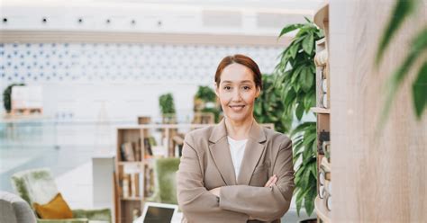 성인 미소 갈색 머리 비즈니스 여성 40 년 동안 긴 머리를 세련 된 베이지 색 정장과 청바지에 공공 장소 녹색 열린 공간 사무실 공동 작업 도서관에서 책을 가진