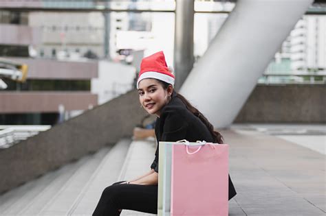 산타 모자와 화려한 쇼핑 가방 크리스마스 선물을 위한 쇼핑 후 야외에서 앉아 웃는 젊은 아시아 여자의 모습 가방에 대한 스톡 사진 및 기타 이미지 Istock