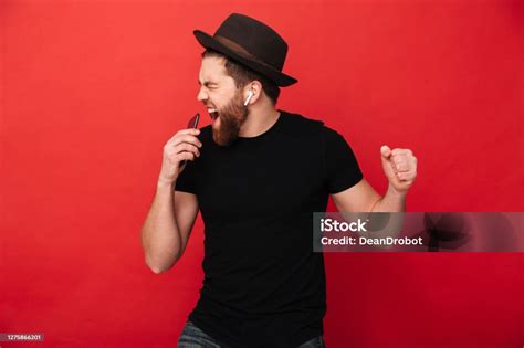 검은 색 티셔츠와 모자를 쓰고 노래하고 빨간 벽 위에 고립 된 휴대 전화와 무선 이어폰으로 음악을 듣는 즐거운 남자의 이미지 T 셔츠에 대한 스톡 사진 및 기타 이미지