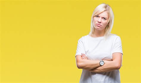 젊은 아름 다운 금발 여자 격리 된 배경 회의론 및 교차 팔과 얼굴에 긴장 Disapproving 식 흰색 T셔츠를 입고 부정적인 사람입니다 T 셔츠에 대한 스톡 사진 및