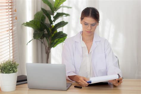 노트북 컴퓨터를 사용하고 클리닉에서 진단을위한 환자에 관한 문서 종이를보고있는 젊은 여성 의사 클립 보드에서 문서를 읽고 일하는