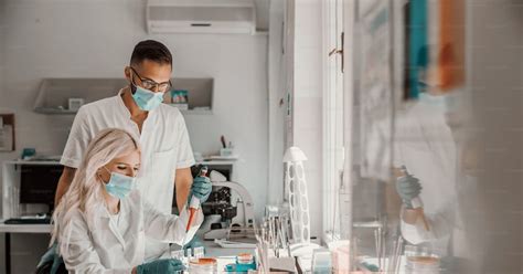 얼굴 마스크와 고무 장갑을 낀 매력적인 헌신적인 금발 여성 실험실 조수가 코로나 바이러스 치료법을 연구하고 있습니다 그녀 옆에는 동료가 서서 그녀를 지켜보고 있습니다