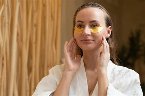 욕실 거울에 서있는 동안 화장품 눈 패치를 적용 목욕 가운을 입고 젊은 여자 가정 생활에 대한 스톡 사진 및 기타 이미지