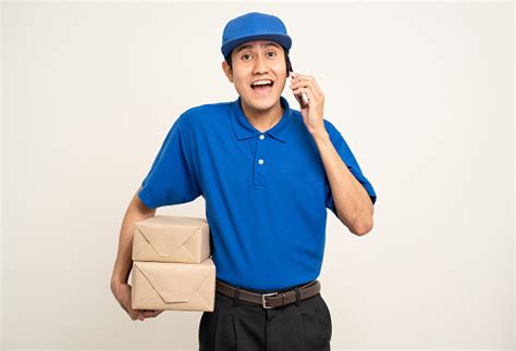 파란색 유니폼을 입은 아시아 남자는 격리된 흰색 배경에 상자 소포 판지와 스마트폰을 들고 서 있습니다 휴대 전화를 가진 남성